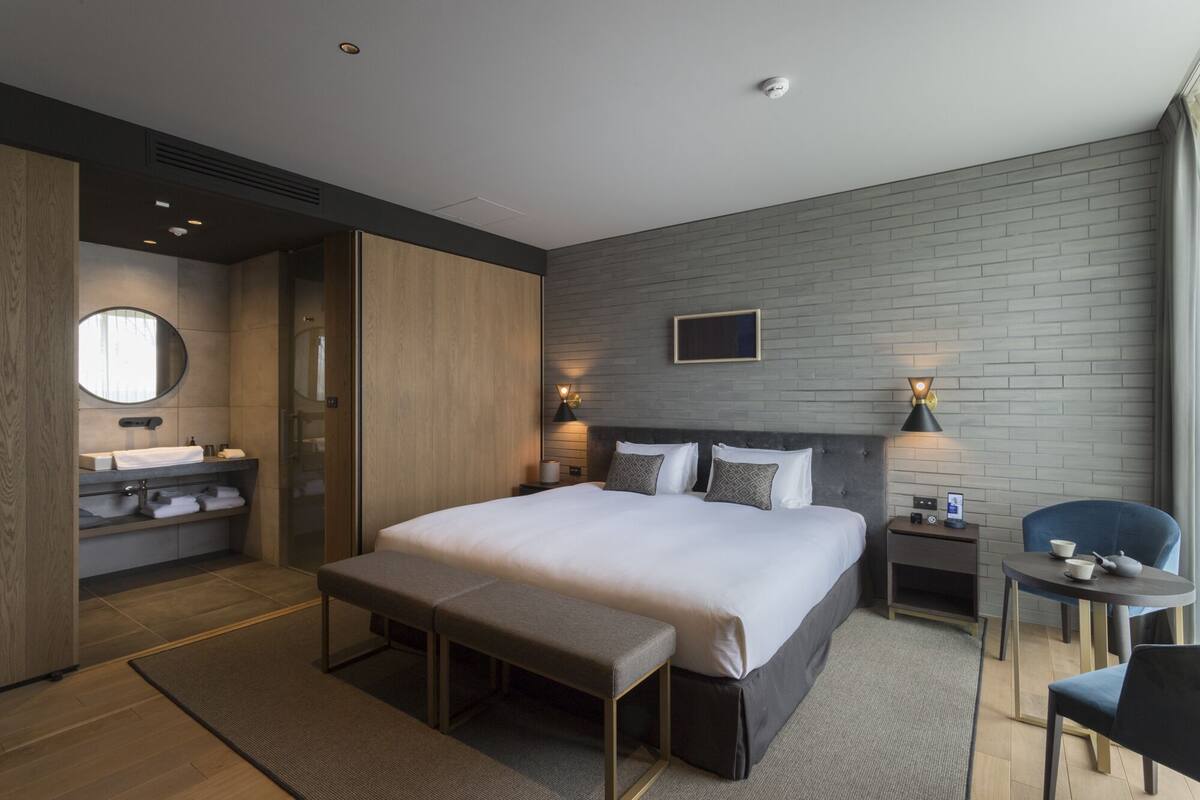Quarto de hotel moderno com uma cama grande, mesas de cabeceira, um espelho redondo, uma pequena mesa com uma cadeira e um banheiro com pia e espelho.