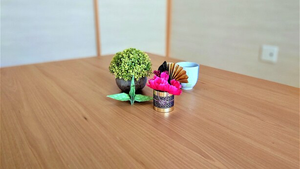 Uma mesa de madeira com uma planta em vaso, um vaso decorativo com flores cor-de-rosa e uma xícara azul.