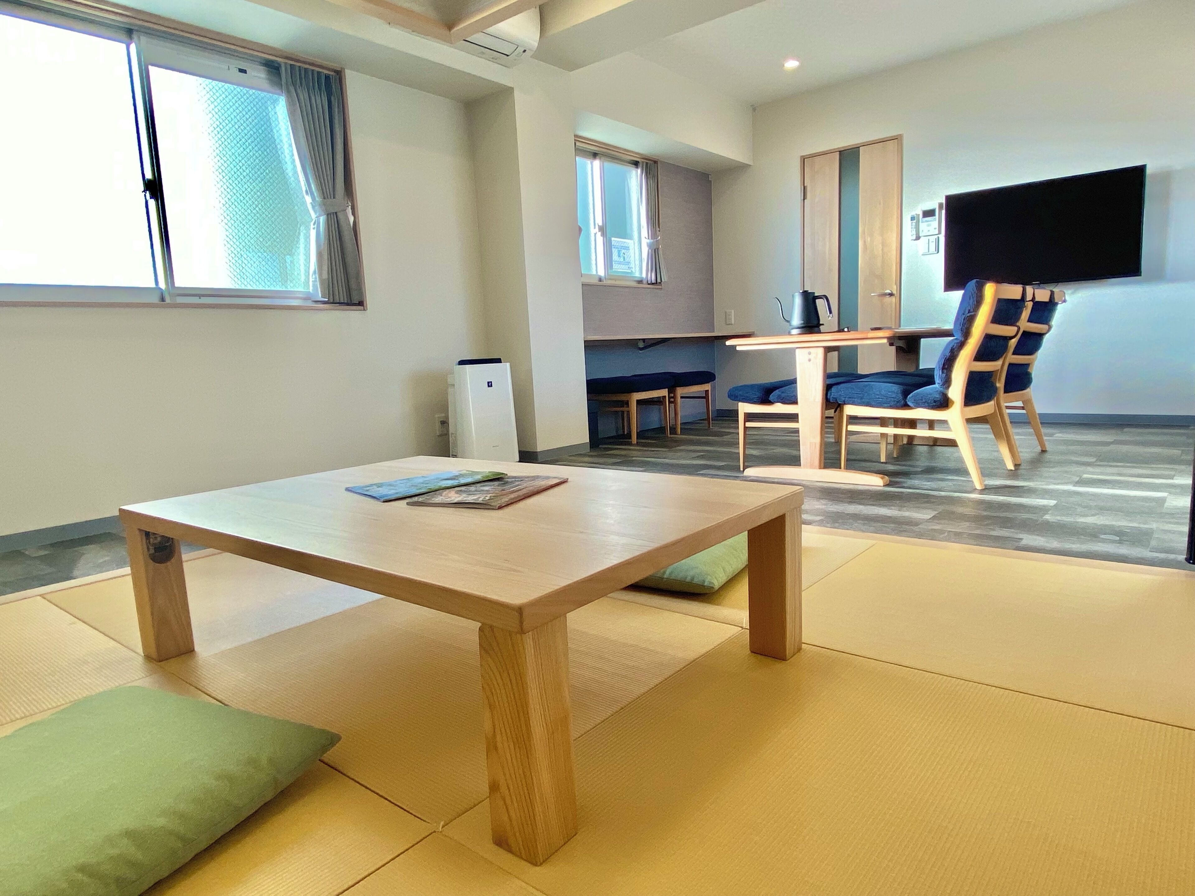 Quarto com tatames, uma mesa de madeira com livros, televisão e área de cozinha compacta.