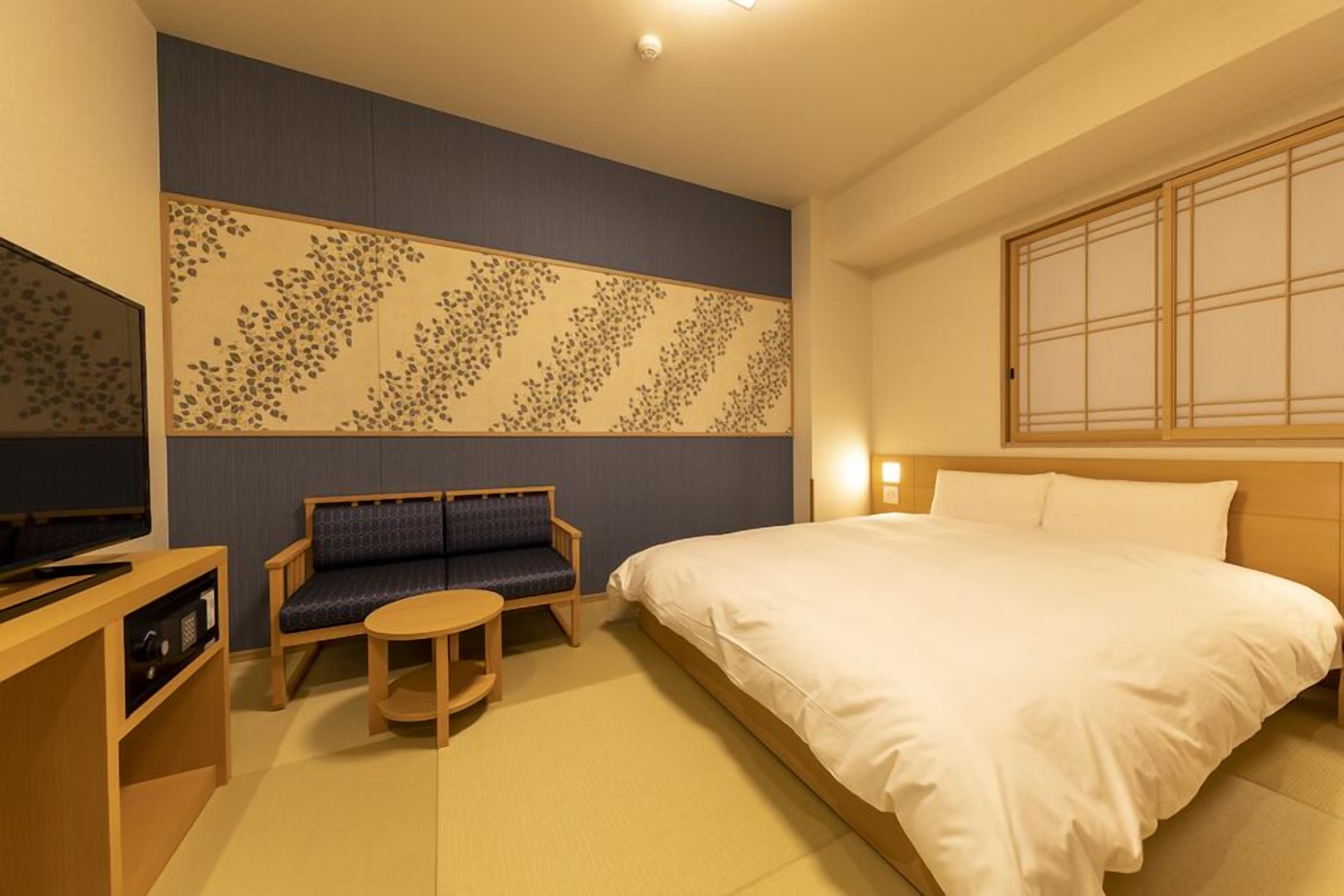 Quarto de hotel com cama, sofá, mesa pequena, televisão e painel decorativo na parede.