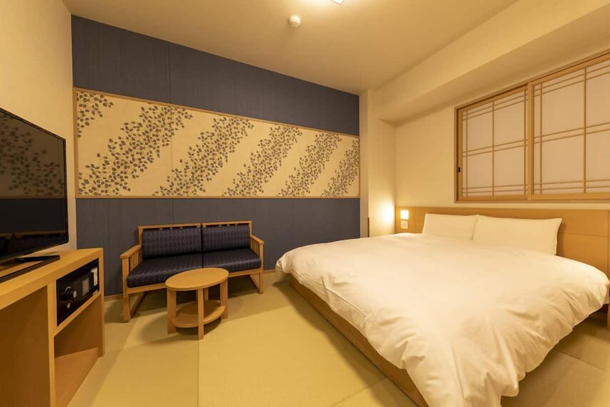Quarto de hotel com cama, sofá, mesa pequena, televisão e painel decorativo na parede.