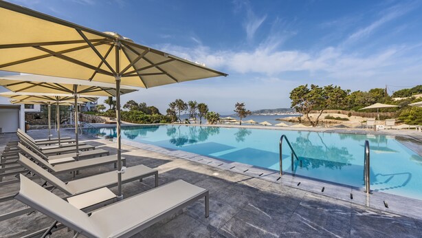 Piscina com espreguiçadeiras e guarda-sóis, rodeada por um jardim com árvores e vista para o mar.