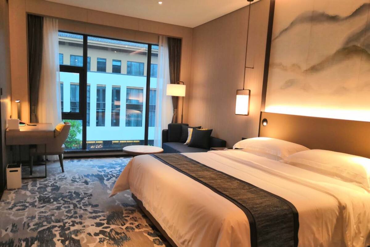 Quarto de hotel com uma cama grande, uma escrivaninha com cadeira, uma mesa redonda pequena e vista para um edifício pela janela.