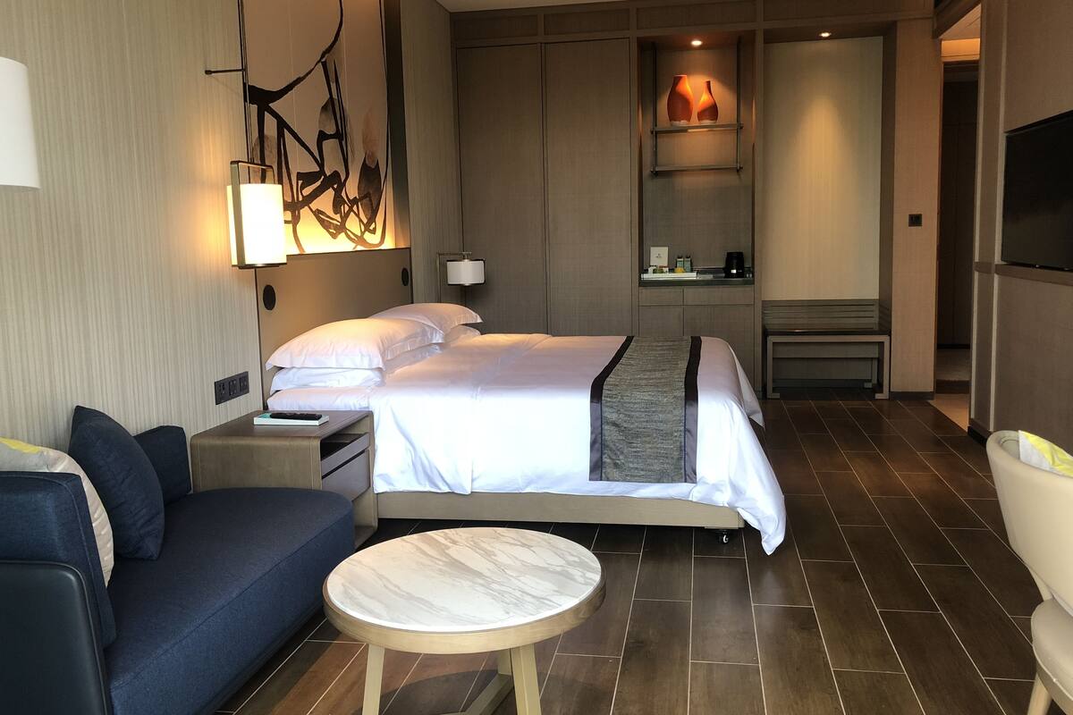 Quarto de hotel com cama, mesa redonda, sofá, televisão e pequena área de cozinha.