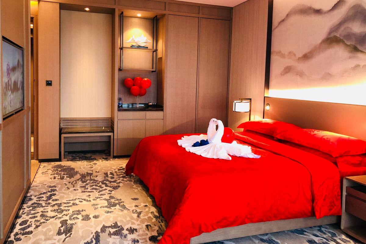 Quarto de hotel com uma cama grande, uma escrivaninha pequena, uma prateleira na parede com balões e um quadro na parede.