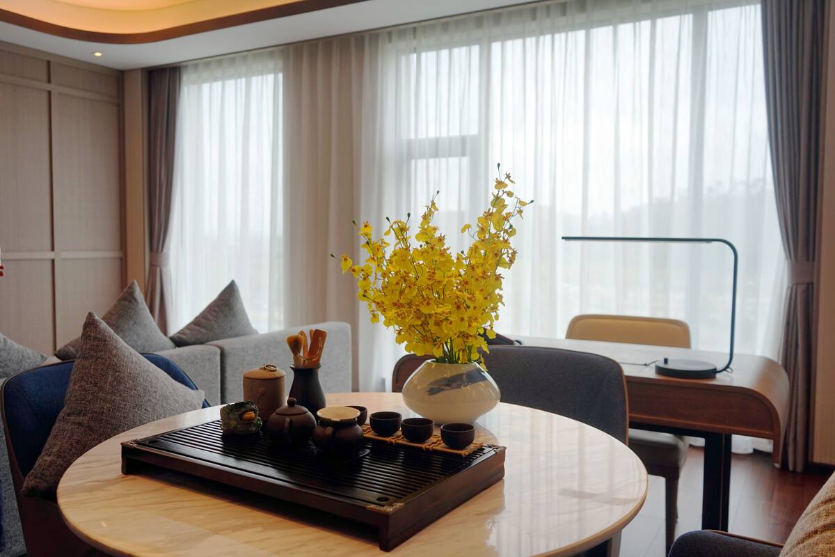 Quarto de hotel moderno com uma mesa de jantar redonda, um conjunto de sofás, um vaso com flores amarelas e um conjunto de chá.