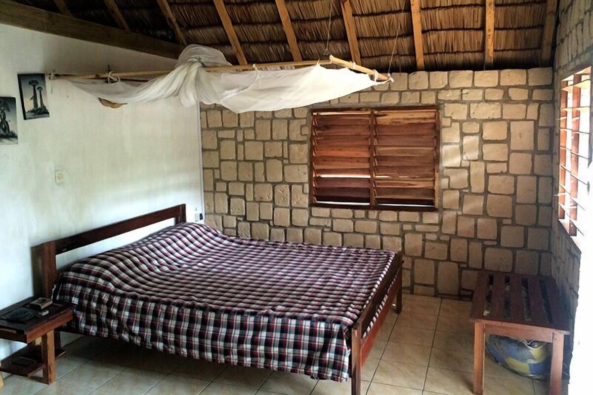 Um quarto com uma cama, um banco de madeira, uma parede de pedra, uma janela com persianas e um telhado de palha.