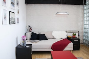 Uma sala de estar moderna com um sofá branco, almofadas preto e branco, um cobertor vermelho, uma mesinha de apoio com uma planta e um candeeiro suspenso.