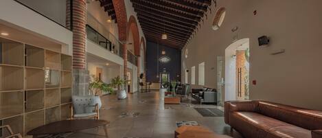 Um hall amplo com pé direito alto, arcos de tijolo aparente e diversos espaços de estar, incluindo um sofá de couro e uma mesa de madeira.