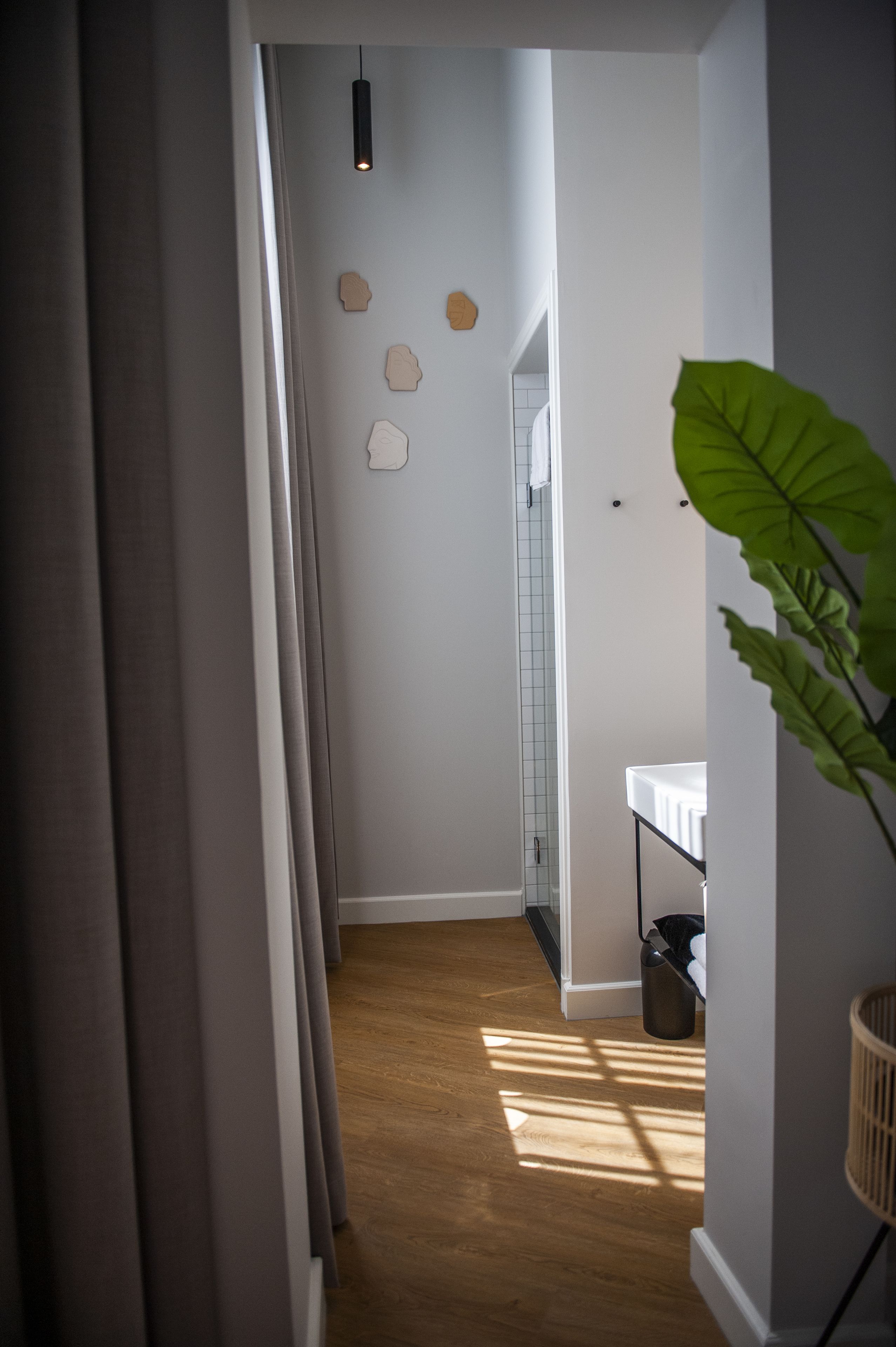 Um corredor estreito com piso de madeira, parede branca com detalhes circulares, luminária pendente preta e um vaso com planta.