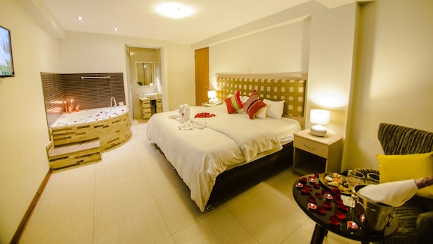 Quarto de hotel com uma cama, uma mesa de cabeceira com abajur, uma pequena mesa com bandeja de frutas e uma xícara, e vista para a área da piscina.