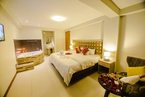 Quarto de hotel com uma cama, uma mesa de cabeceira com abajur, uma pequena mesa com bandeja de frutas e uma xícara, e vista para a área da piscina.