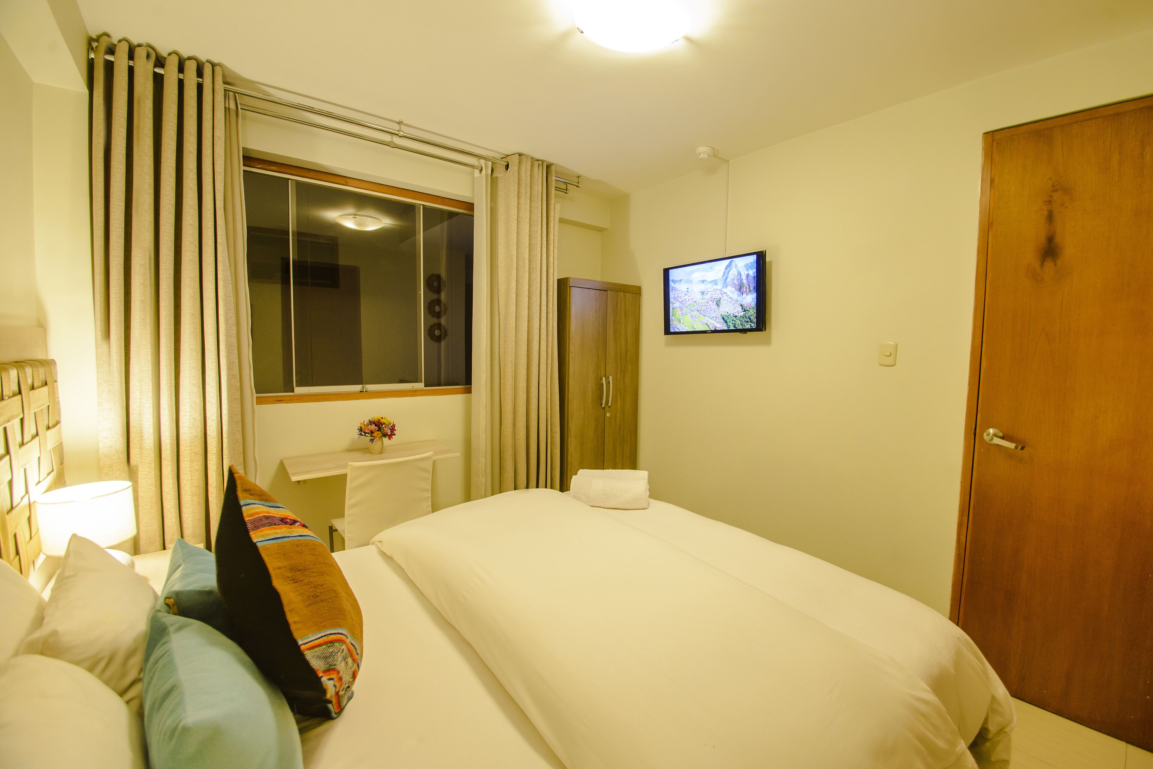 Quarto de hotel com cama, escrivaninha, televisão e guarda-roupa.