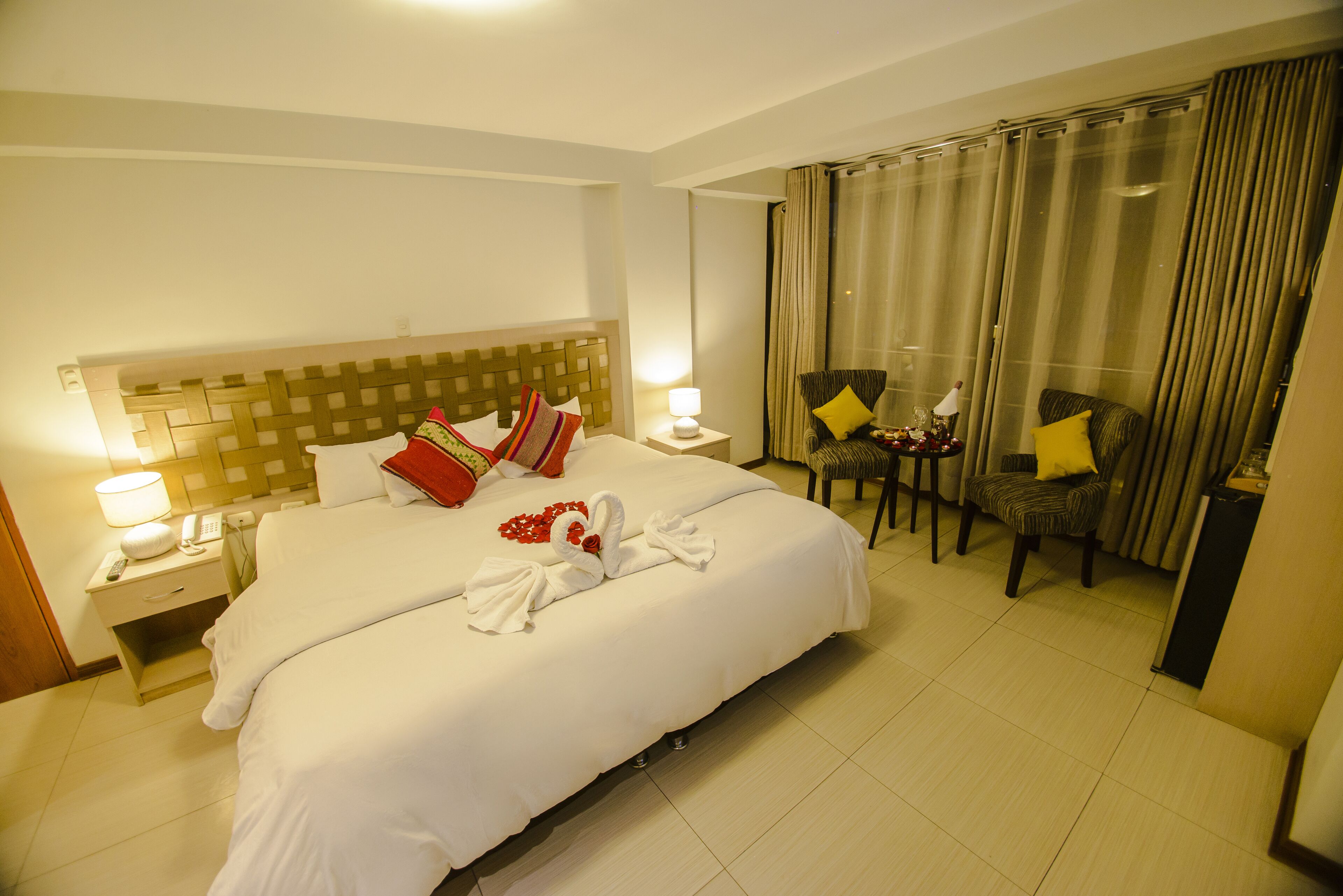 Quarto de hotel com uma cama grande, uma mesa de cabeceira com abajur, uma cadeira e uma pequena mesa com luminária.