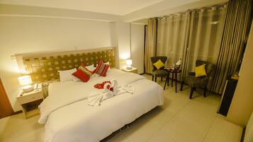Quarto de hotel com uma cama grande, uma mesa de cabeceira com abajur, uma cadeira e uma pequena mesa com luminária.