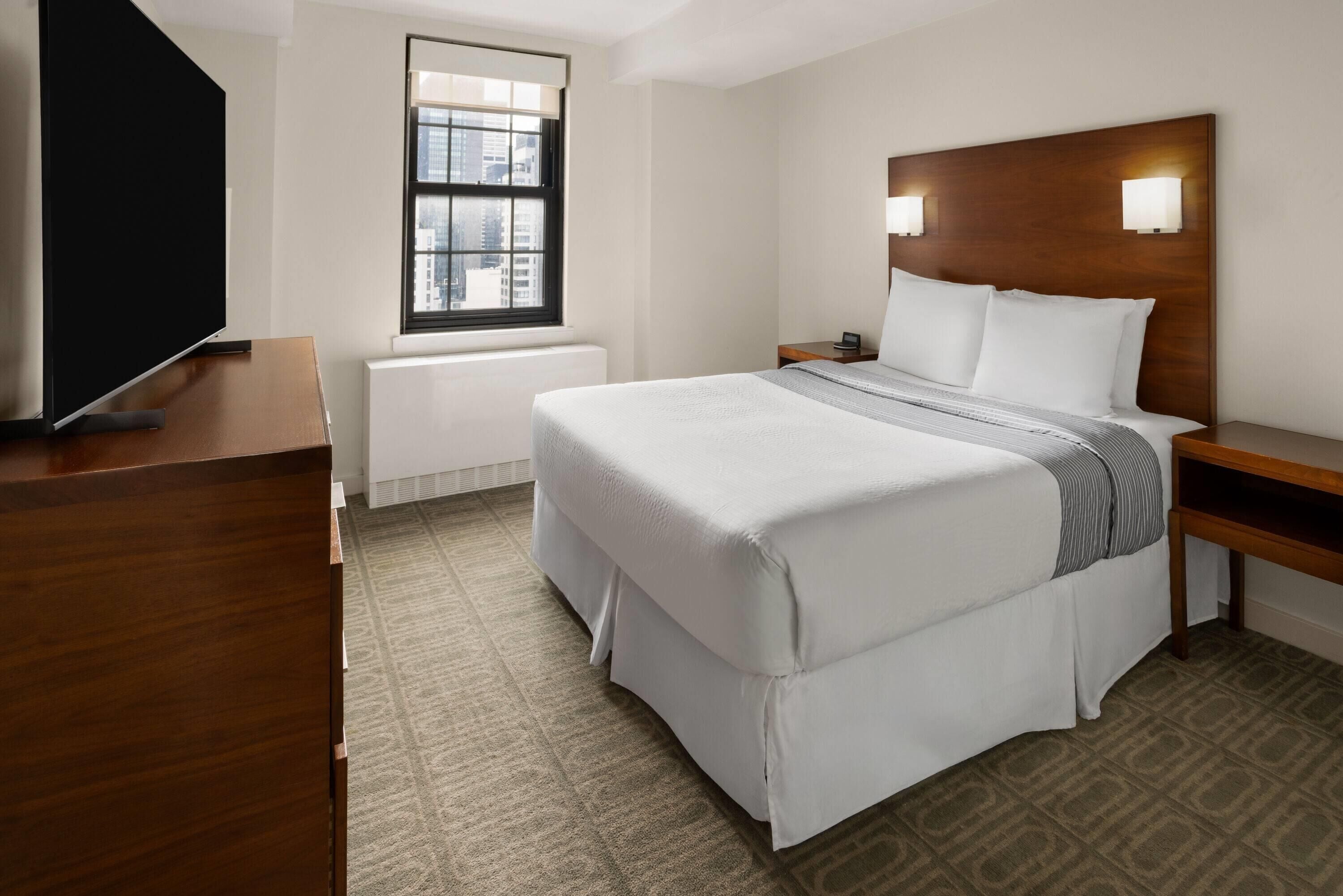 Quarto de hotel com uma cama grande, uma TV sobre uma escrivaninha de madeira, uma janela com vista para a cidade e uma cabeceira com luzes fixadas na parede.