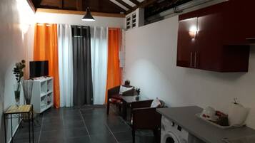Cozinha compacta com máquina de lavar, área de refeições com mesa e cadeiras, e uma TV sobre um suporte.