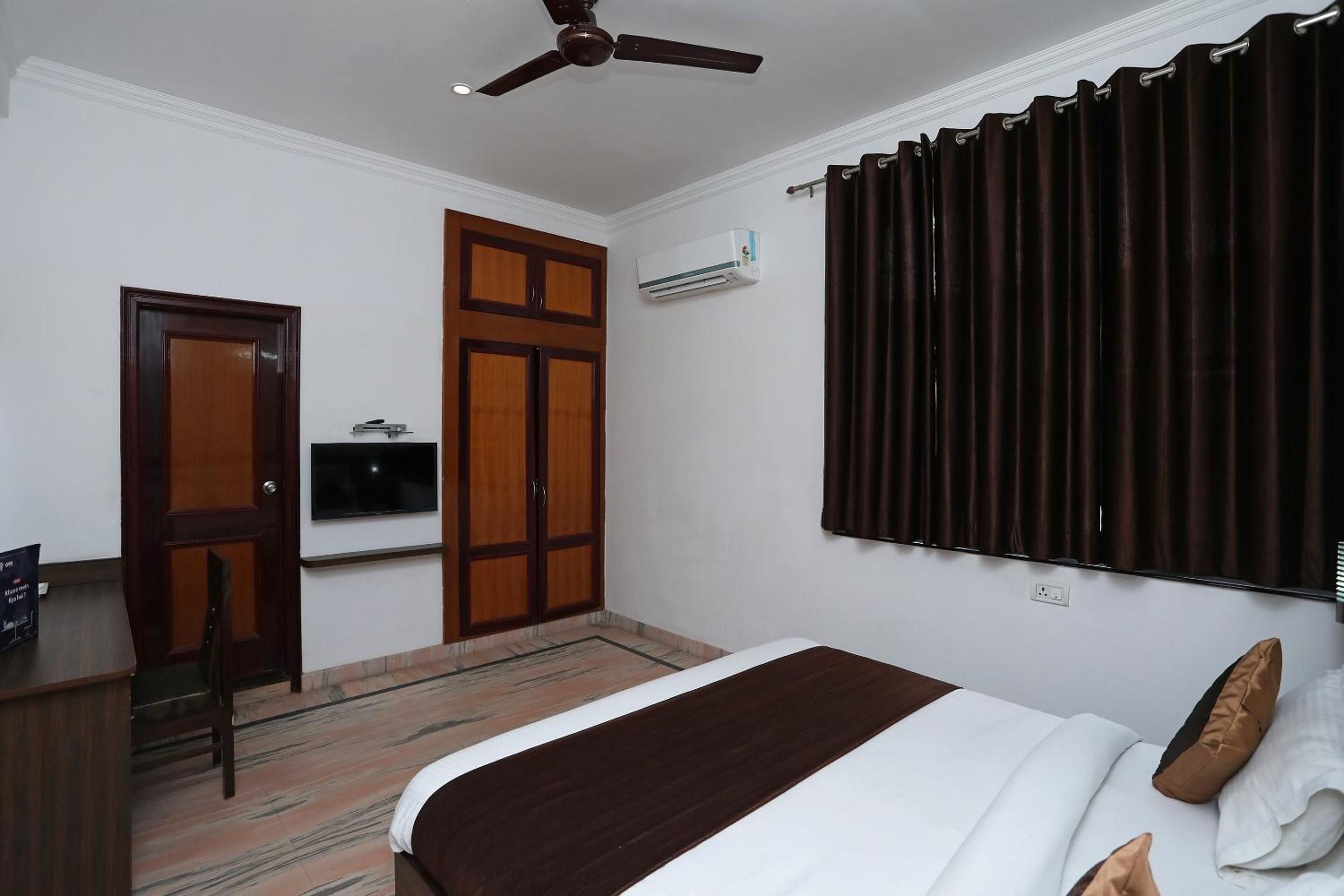 Quarto de hotel com cama, escrivaninha, televisão, aparelho de ar condicionado e ventoinha de teto.