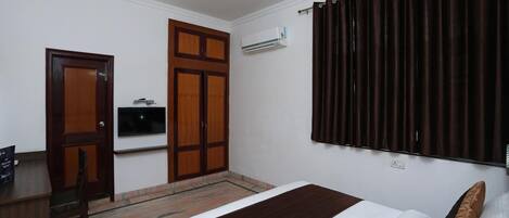 Quarto de hotel com cama, escrivaninha, televisão, aparelho de ar condicionado e ventoinha de teto.