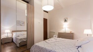 Quarto de hotel com um espelho grande, guarda-roupa de madeira, luminárias de cabeceira, uma cama com colcha estampada e uma pequena mesa de cabeceira com abajur.
