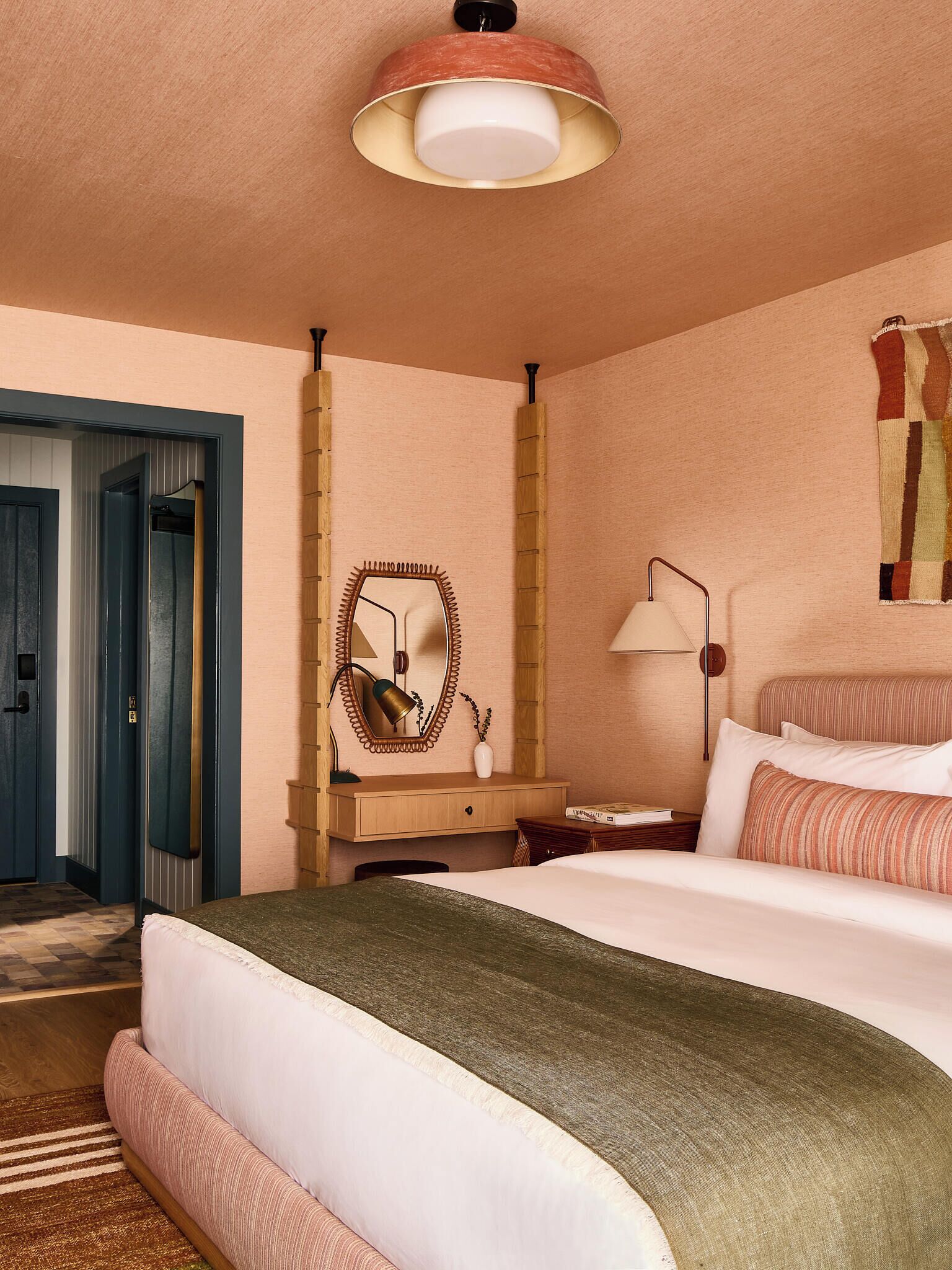 Quarto de hotel com cama, mesa de cabeceira, luminária, espelho e porta.