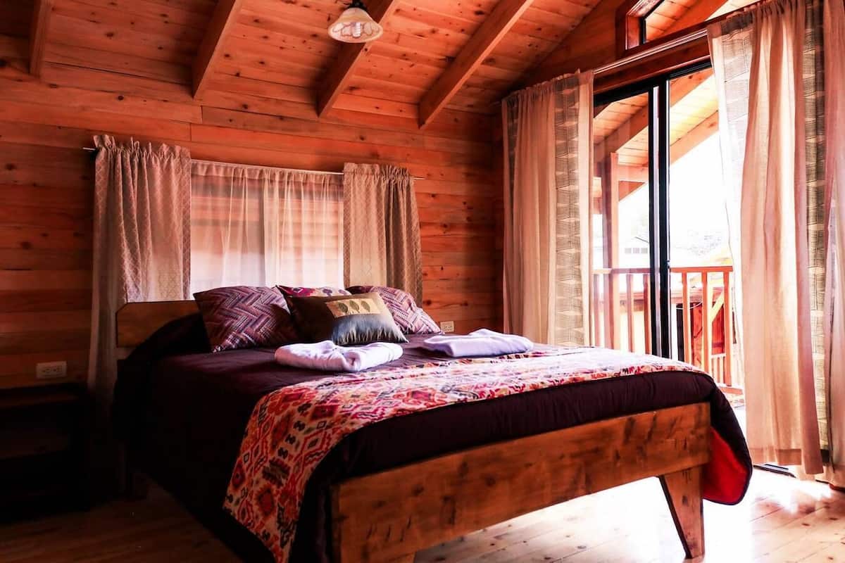 Quarto de cabana de madeira com cama, cortinas e vista para o exterior.