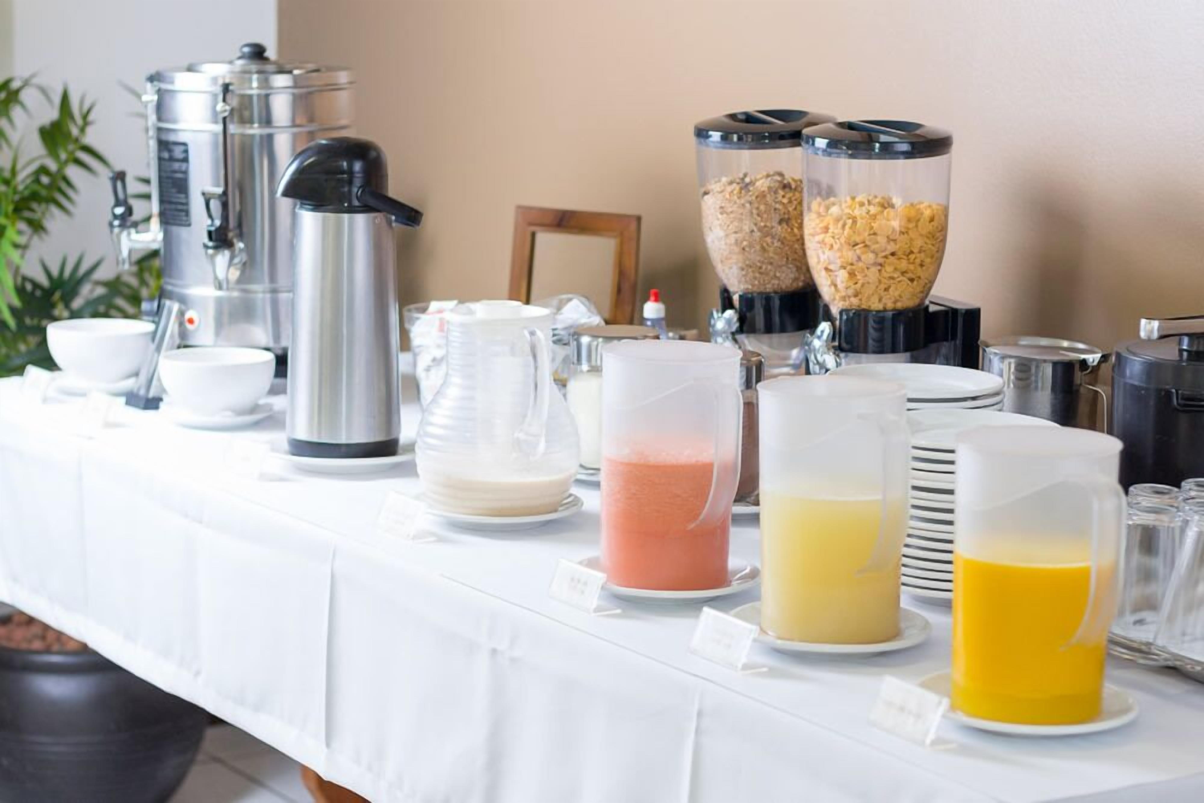 Disposição de buffet com diversos dispensadores de comida e bebida, incluindo uma cafeteira e recipientes para cereais e sumos.