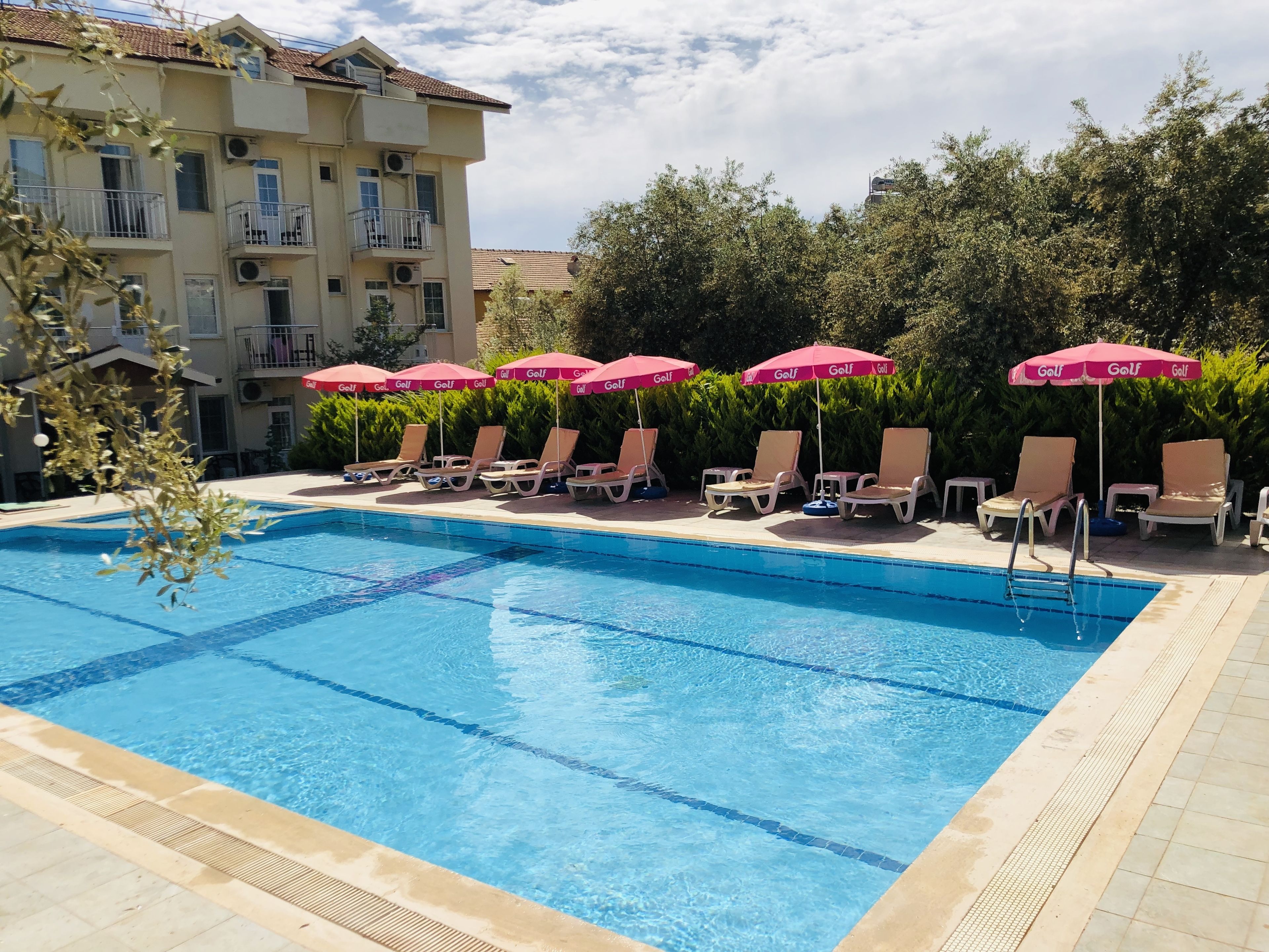 Uma piscina com espreguiçadeiras e guarda-sóis, rodeada por vegetação e um edifício com varandas.
