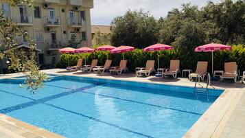 Uma piscina com espreguiçadeiras e guarda-sóis, rodeada por vegetação e um edifício com varandas.