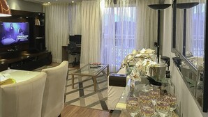 Uma sala de estar moderna com uma TV de tela plana, um sofá, uma mesa de centro e uma vitrine de vidro com objetos decorativos.
