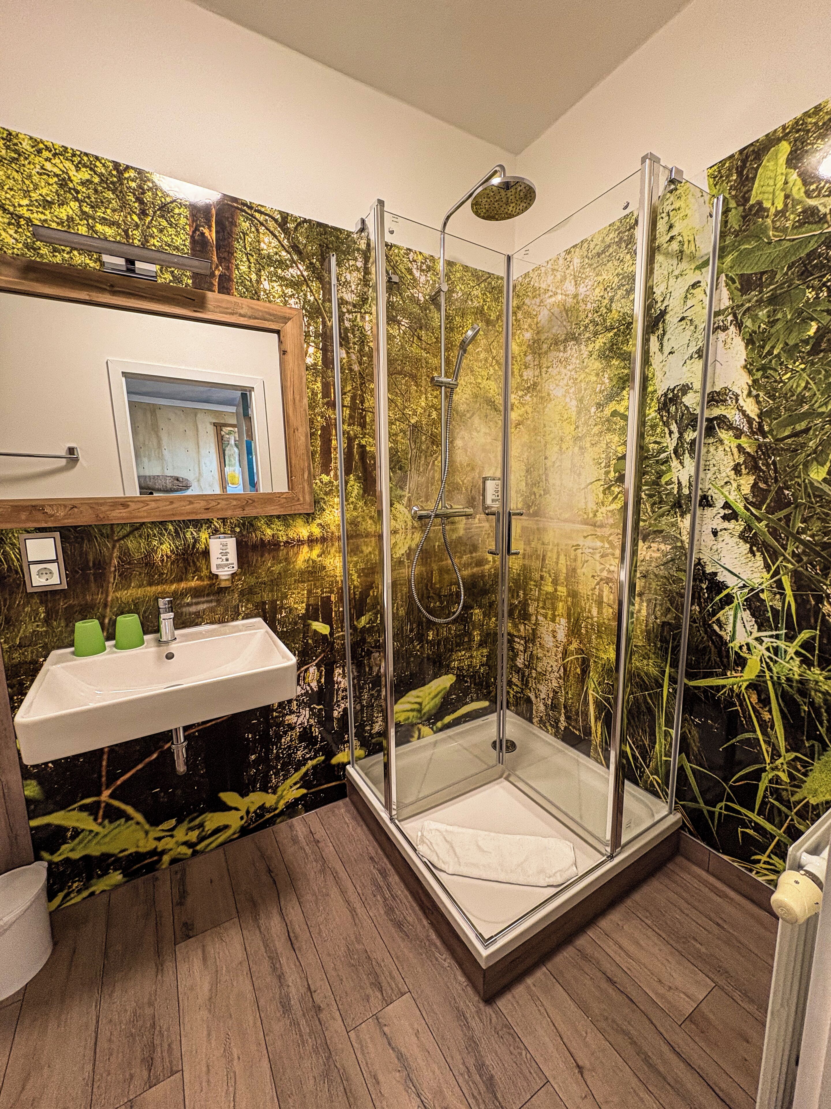 Casa de banho com cabine de duche em vidro, lavatório com espelho e painel mural com tema de floresta.
