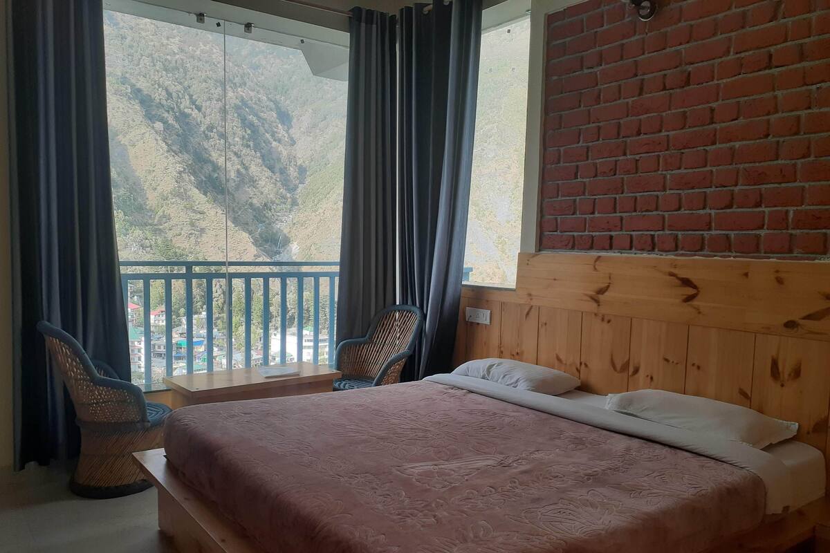 Quarto de hotel com uma cama grande, cabeceira de madeira, vista para uma paisagem montanhosa e varanda com uma cadeira.