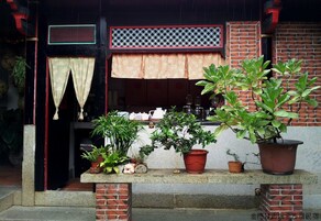 Fachada de tijolos com uma pequena varanda. Há plantas em vasos na varanda, uma janela com cortina e um painel decorativo acima da janela.