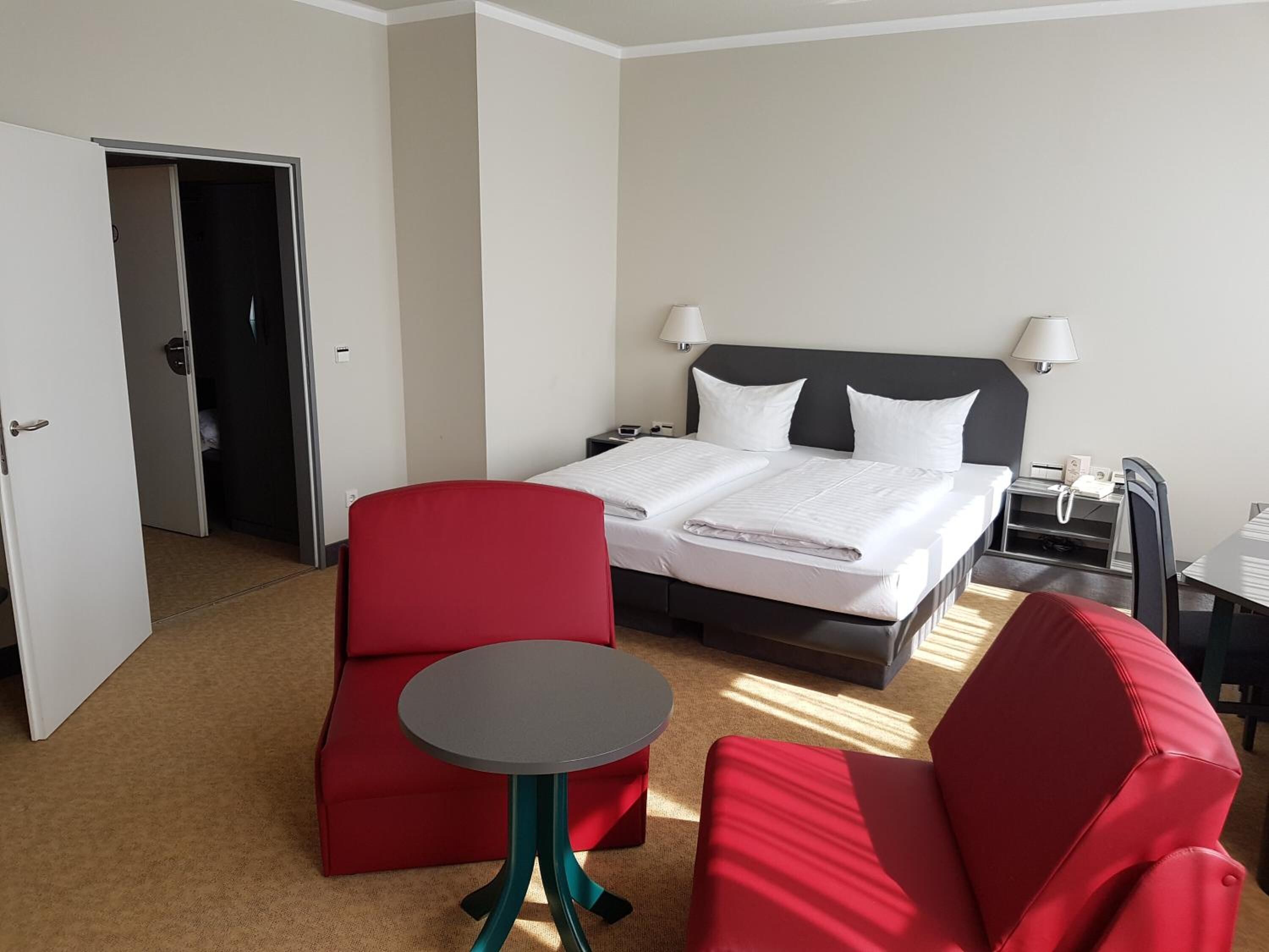 Quarto de hotel com cama, duas cadeiras vermelhas, uma mesinha redonda, mesa de cabeceira com abajur e escrivaninha com cadeira.