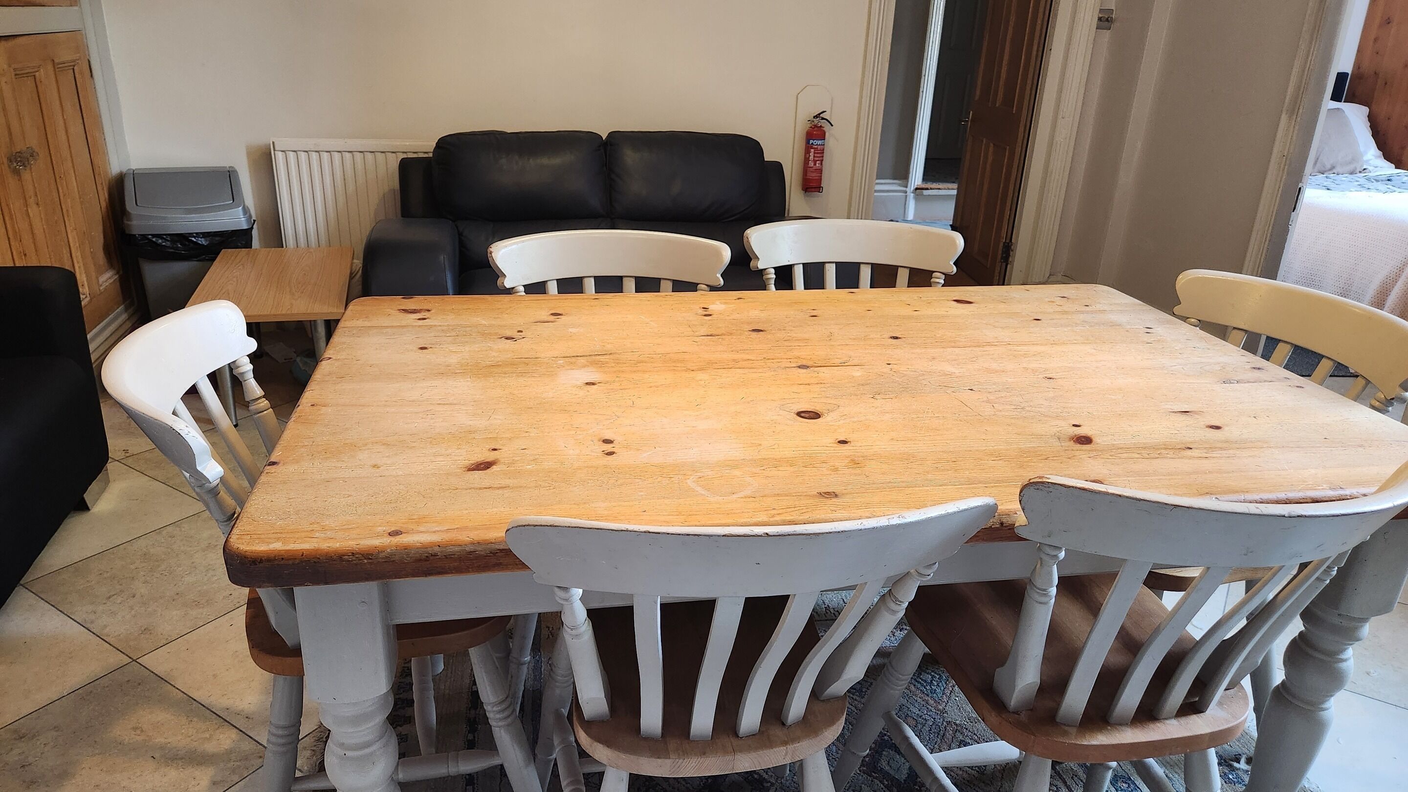 Uma mesa de jantar com seis cadeiras brancas, um sofá preto e um extintor na parede.