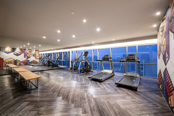 Uma academia com equipamentos de ginástica, piso de madeira e janelas amplas com vista para a cidade.