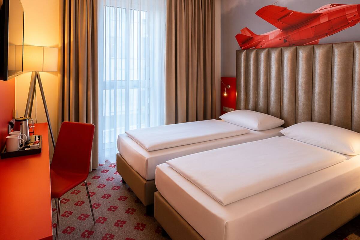 Quarto de hotel com duas camas, uma escrivaninha com cadeira, uma lâmpada, uma decoração de avião vermelha e uma janela com cortinas.