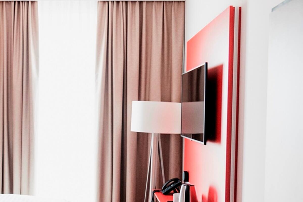 Quarto de hotel com cama, escrivaninha com telefone, luminária e TV na parede.