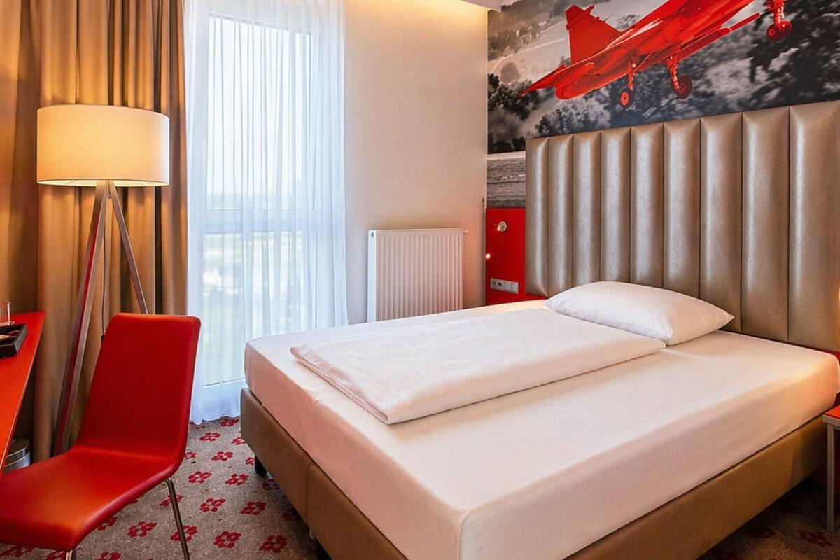 Quarto de hotel com cama, escrivaninha, cadeira, janela com cortinas, radiador e um mural na parede com um avião.