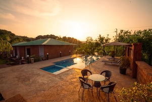 Piscina com área de terraço, incluindo mesa e cadeiras, rodeada por um edifício de tijolos e vegetação.