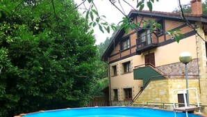 Uma piscina com deck de madeira, cercada por vegetação exuberante e um prédio de dois andares com varanda.