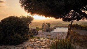 Uma trilha de pedra leva a uma ponte rústica sobre um riacho, emoldurada por vegetação e com vista para o pôr do sol.
