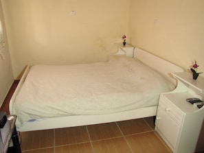 Quarto pequeno com cama de solteiro, colcha branca, mesa de cabeceira com um vaso pequeno e cômoda branca com um controle remoto em cima.
