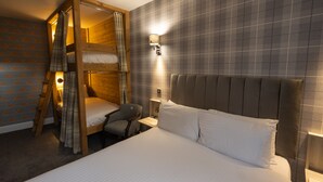 Quarto de hotel com uma cama de solteiro, uma cama individual, uma cadeira, uma mesinha de cabeceira e uma luminária fixada na parede.