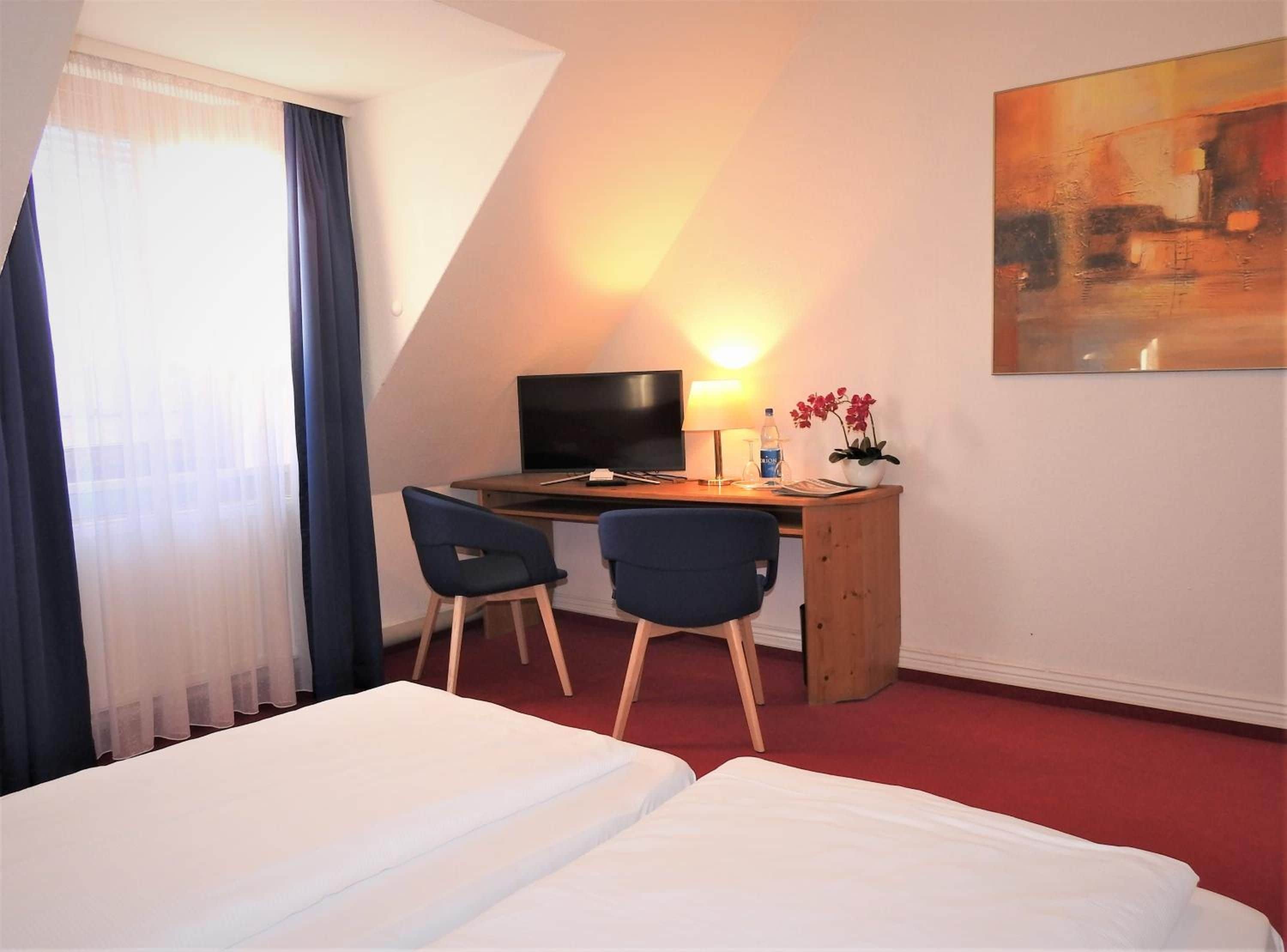 Quarto de hotel com uma cama, uma escrivaninha com duas cadeiras, uma televisão e um quadro na parede.