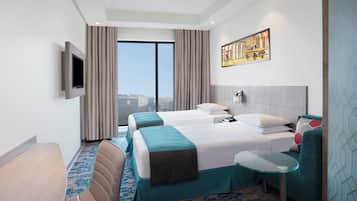 Quarto de hotel com duas camas, televisão, mesinha, sofá e uma janela grande com cortinas.