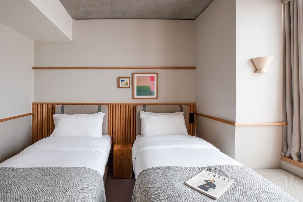 Quarto de hotel com duas camas, cabeceira de madeira, mesa de cabeceira pequena, quadro emoldurado na parede e janela com cortinas.