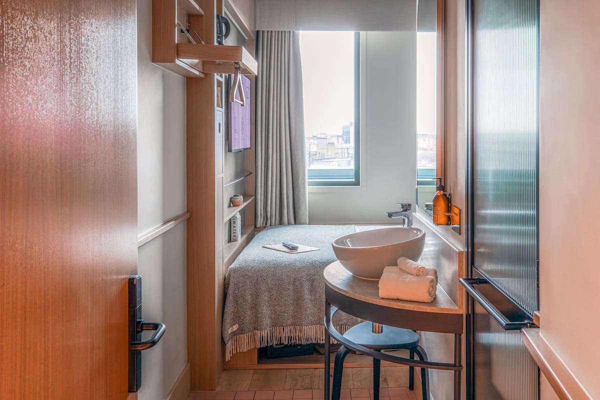 Quarto de hotel compacto com uma janela grande, uma mesa redonda com uma tigela, uma cadeira, uma cama com um cobertor e um guarda-roupa com prateleiras.