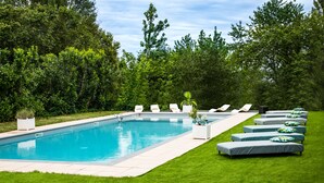 Uma piscina retangular com borda azulejada, cercada por um gramado bem cuidado e árvores verdes exuberantes.