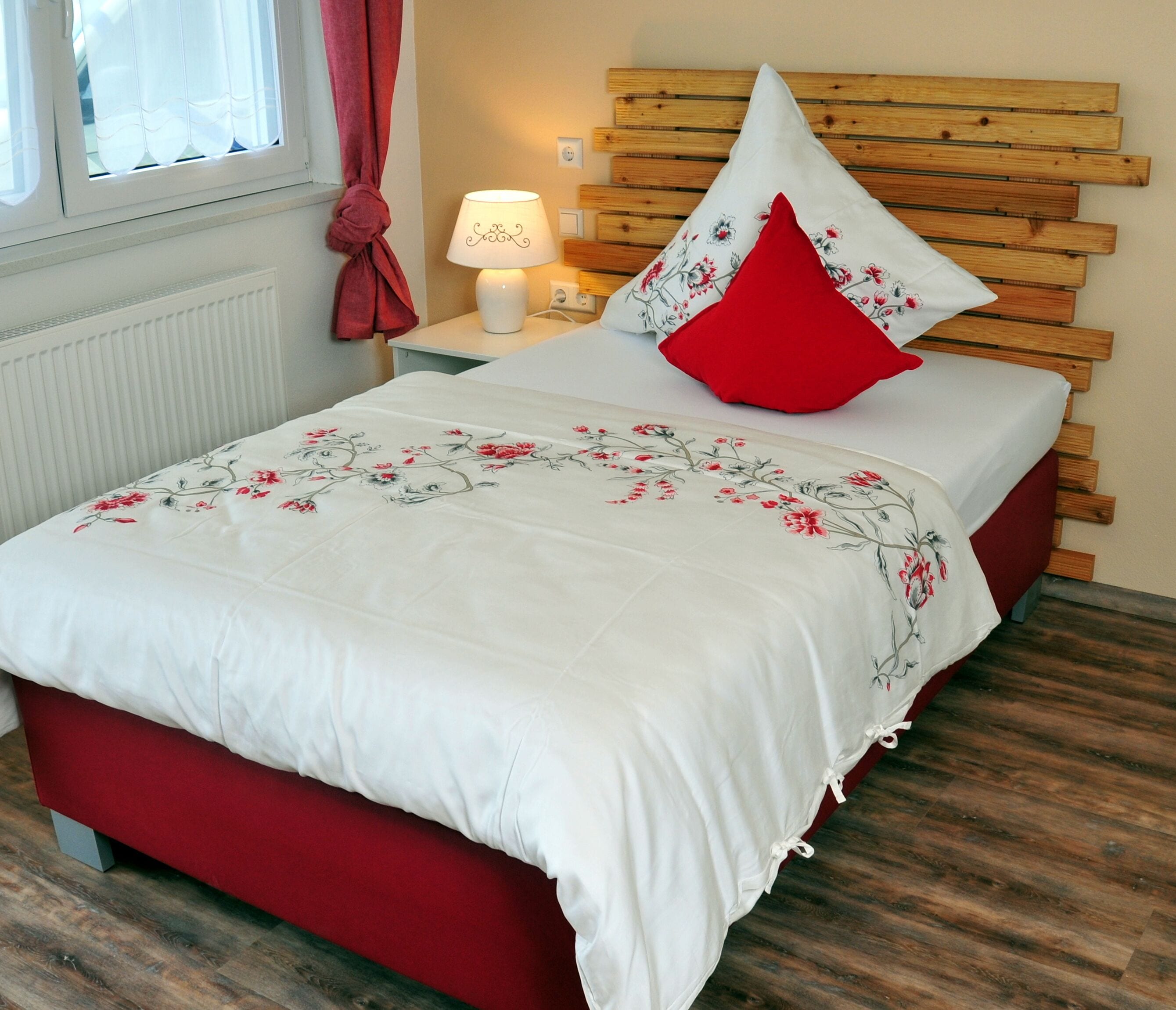 Uma cama com cabeceira vermelha, um edredom branco com motivos florais e um travesseiro vermelho.